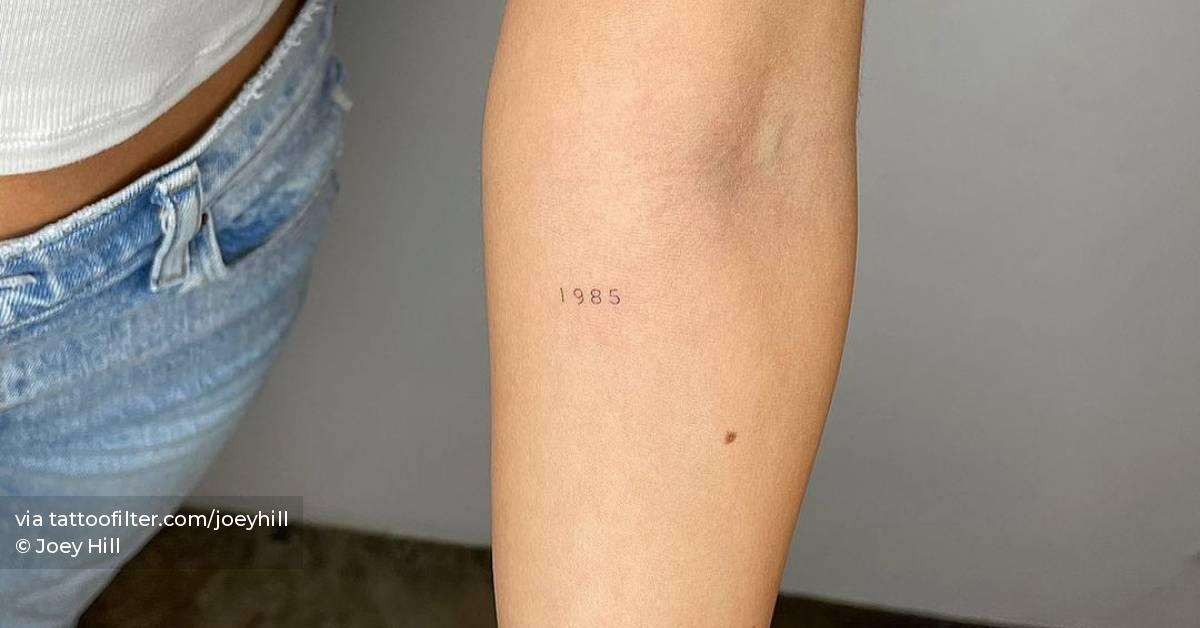 "1985" lettering tattoo on the inner forearm.