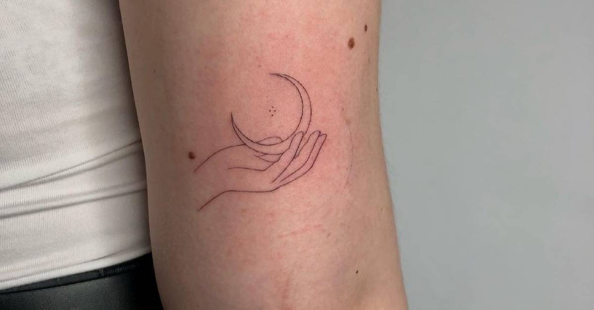 Fine line hand holding moon tattoo on the tricep.