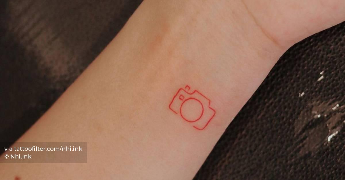 Tiny camera tattoo in red ink located on the wrist.