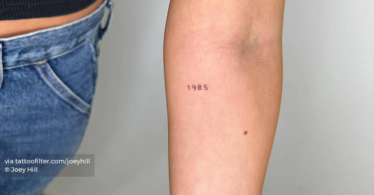 "1985" lettering tattoo placed on the inner forearm.
