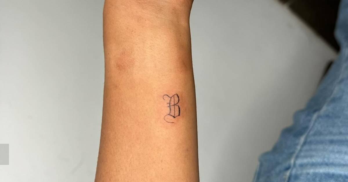 Little tattoo of the letter "B" in gothic font located