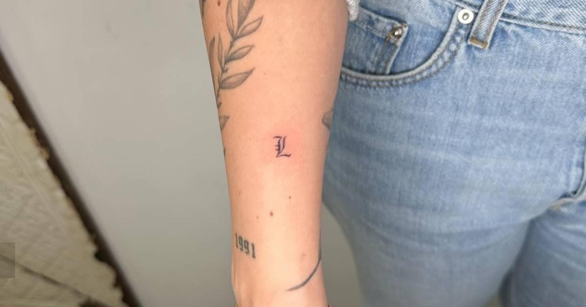 Tiny tattoo of the letter "L" in gothic font located on