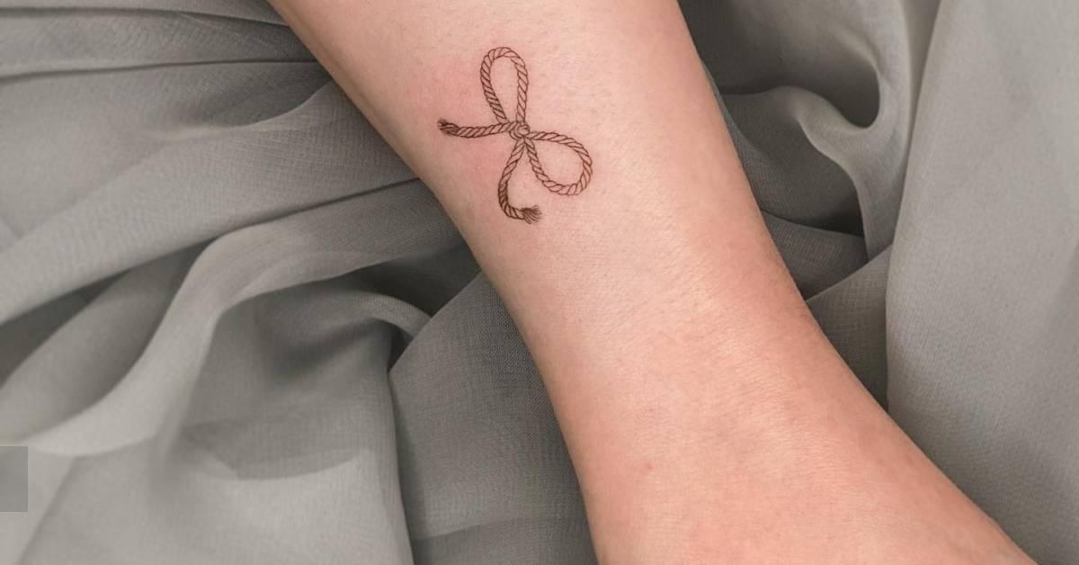 Fine line style rope bow tattoo located on the wrist.